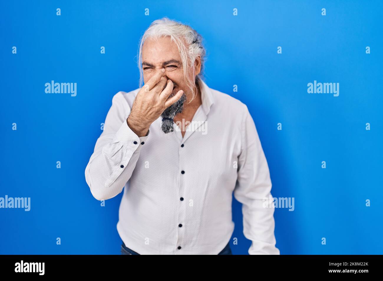 Middle age man with grey hair standing over blue background smelling ...