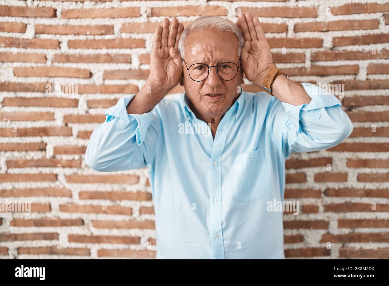 Senior man with grey hair standing over bricks wall doing bunny ears ...