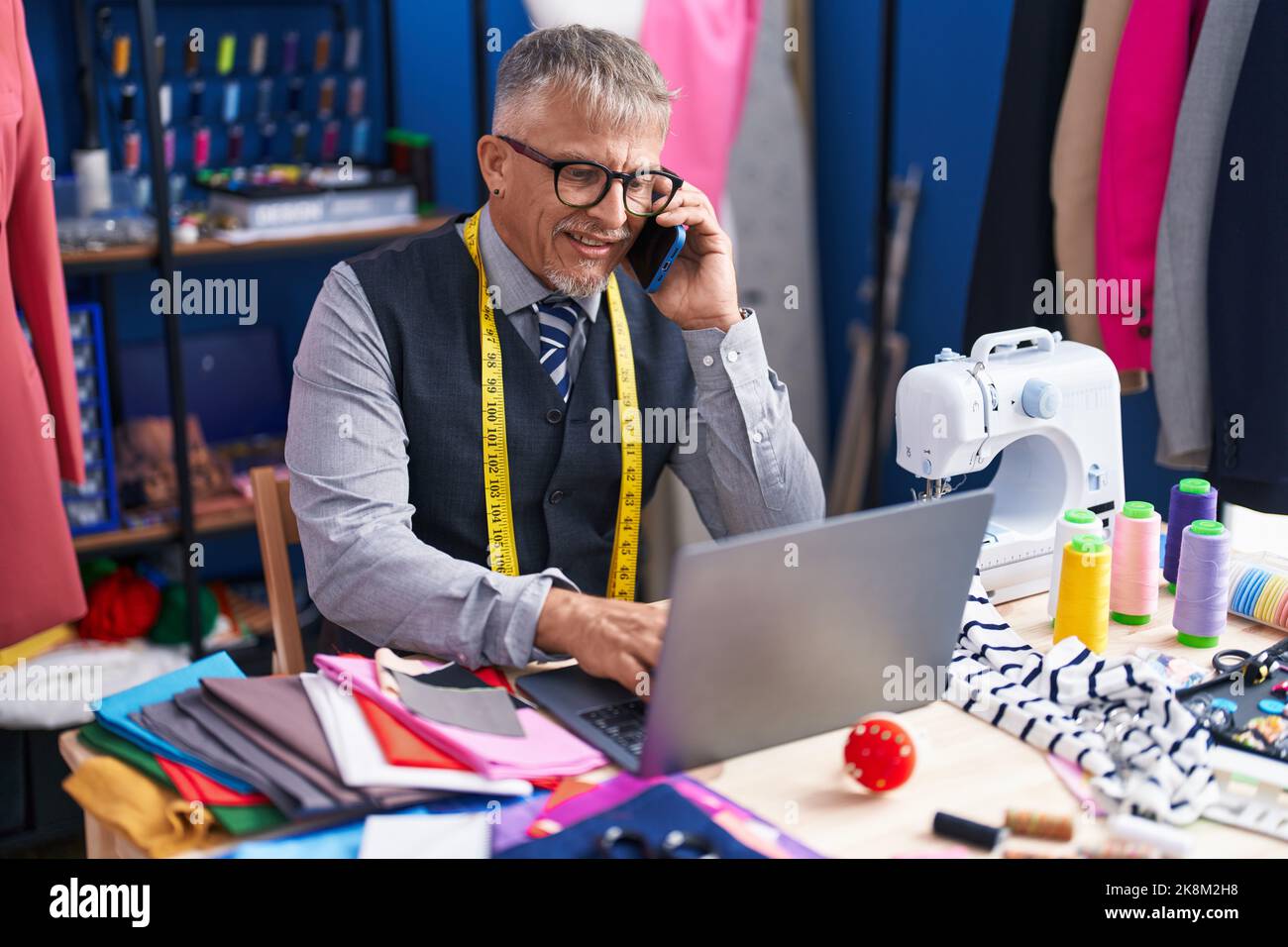 Middle age grey-haired man tailor talking on smartphone using laptop at ...