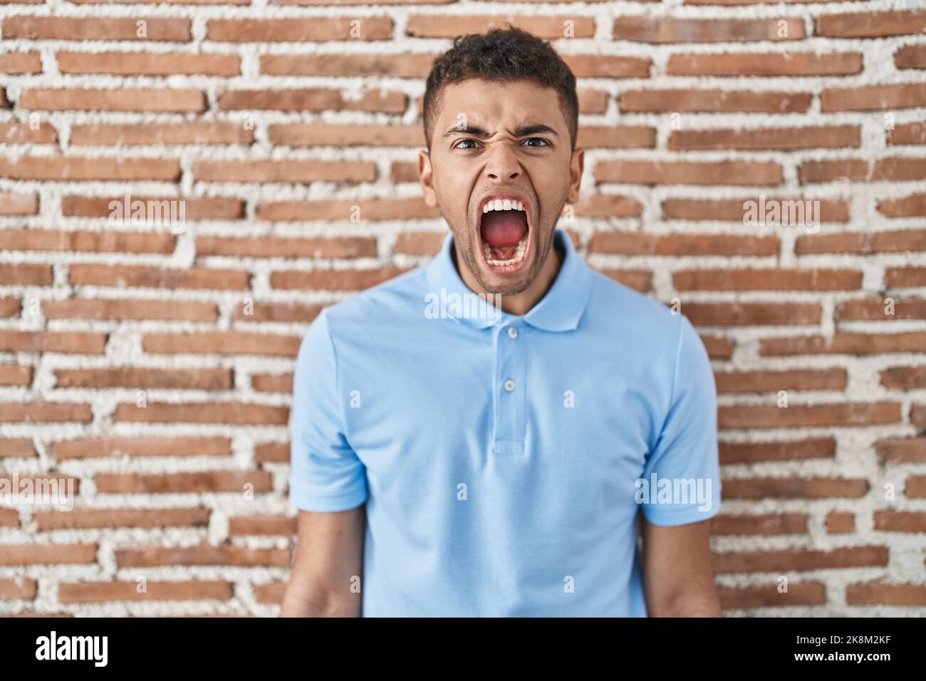 Brazilian young man standing over brick wall angry and mad screaming ...