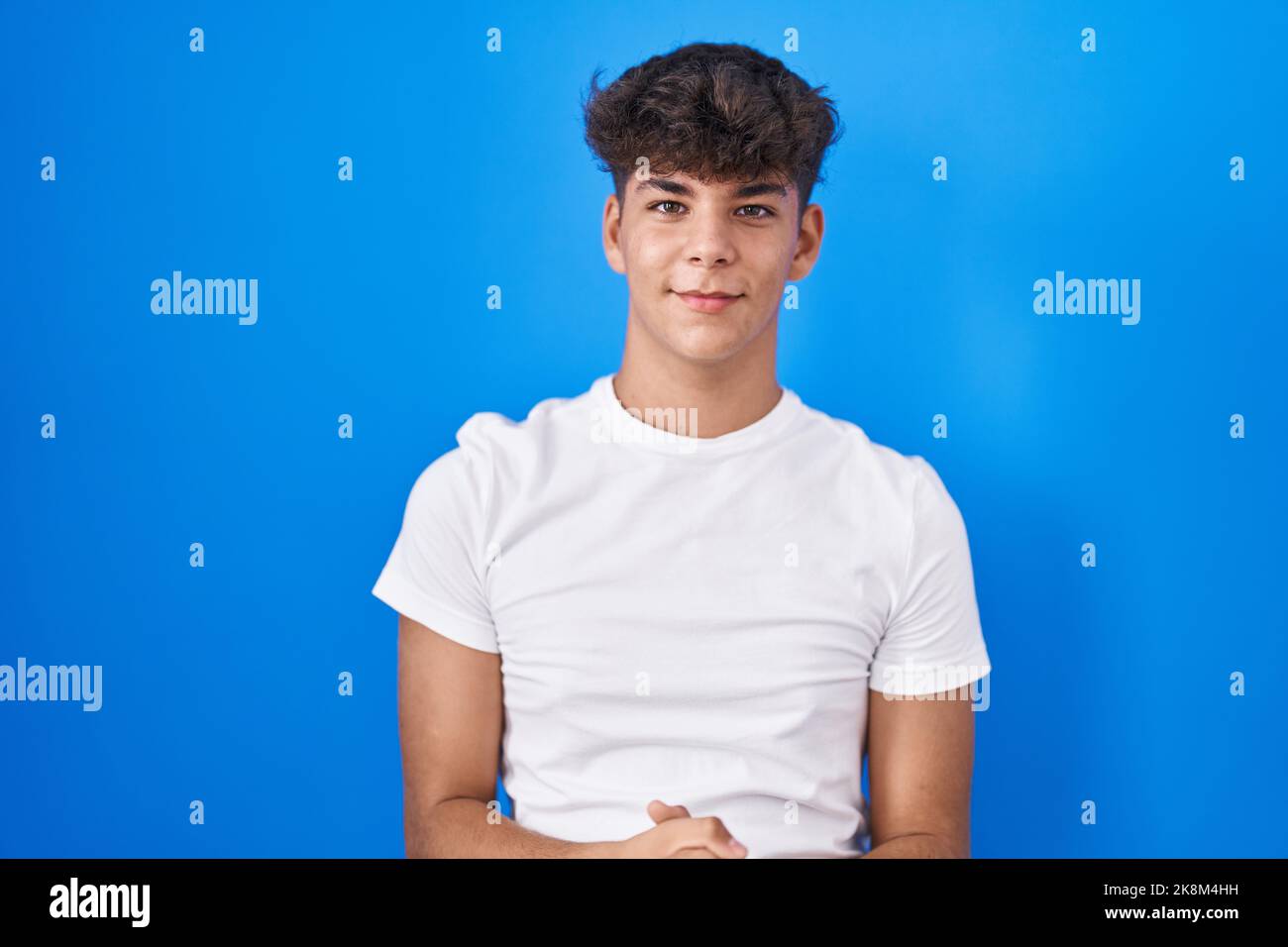 Hispanic teenager standing over blue background with hands together and ...