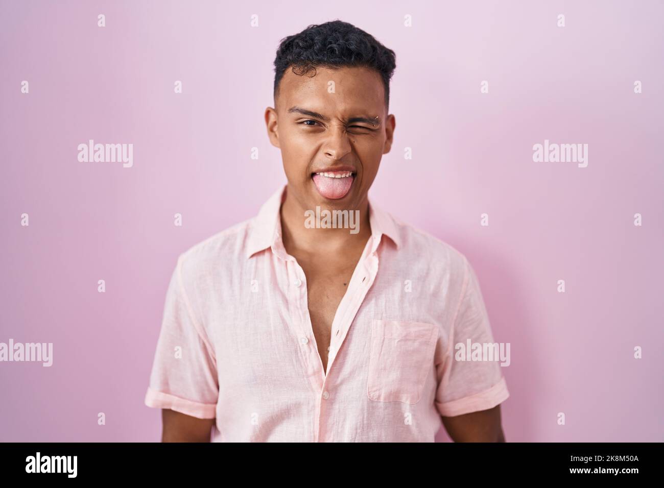 Young hispanic man standing over pink background sticking tongue out ...