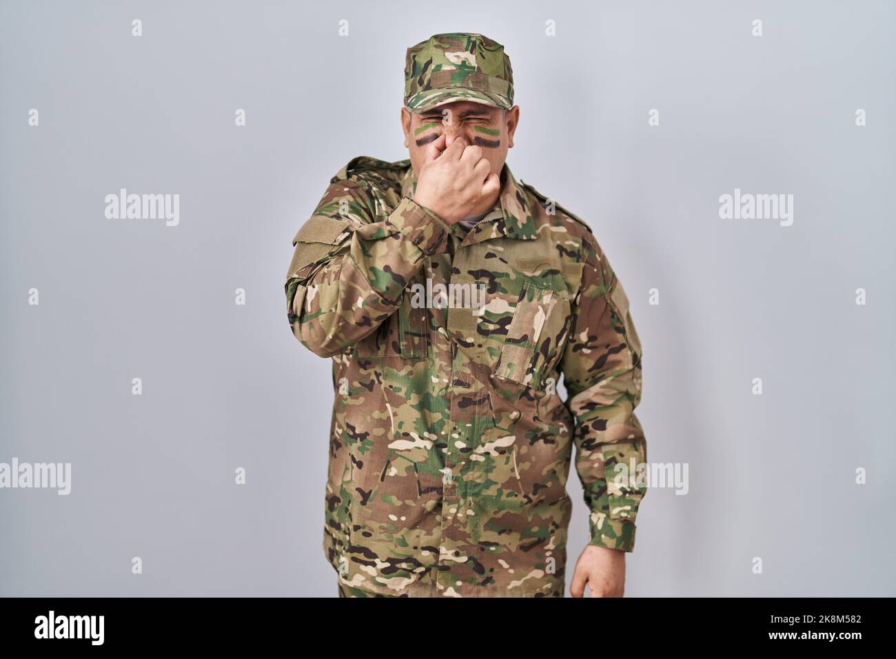 Hispanic young man wearing camouflage army uniform smelling something ...