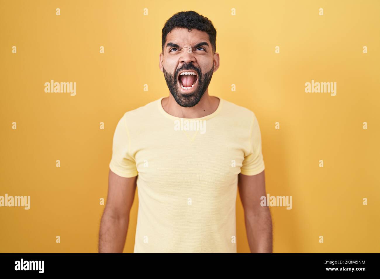 Hispanic man with beard standing over yellow background angry and mad ...