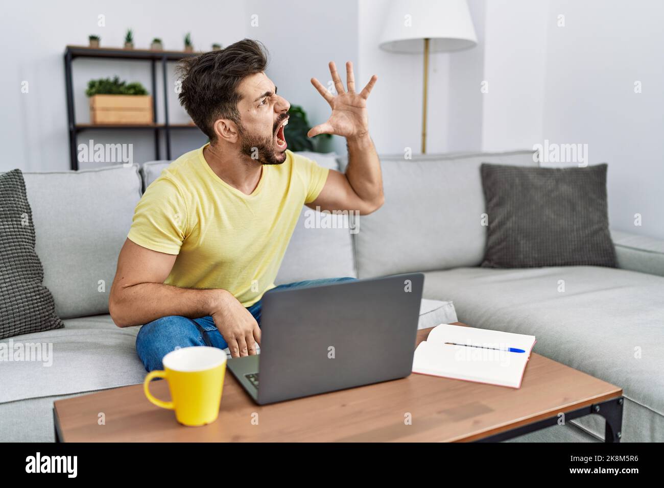 Young man with beard using laptop at home shouting and screaming loud ...