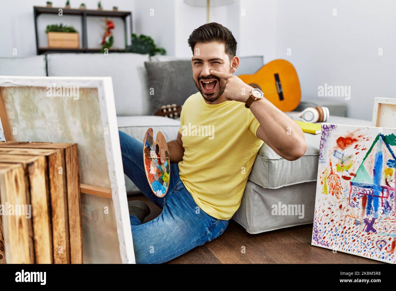 Young man with beard painting canvas at home pointing with hand finger ...
