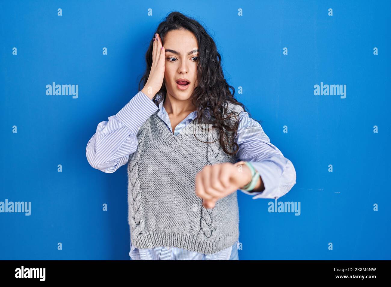 Young brunette woman standing over blue background looking at the watch ...
