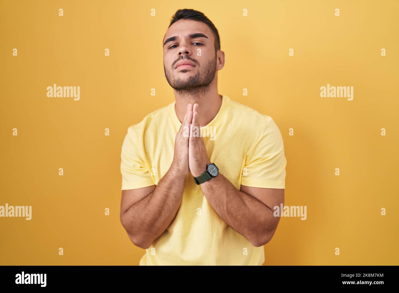 Young hispanic man standing over yellow background begging and praying ...