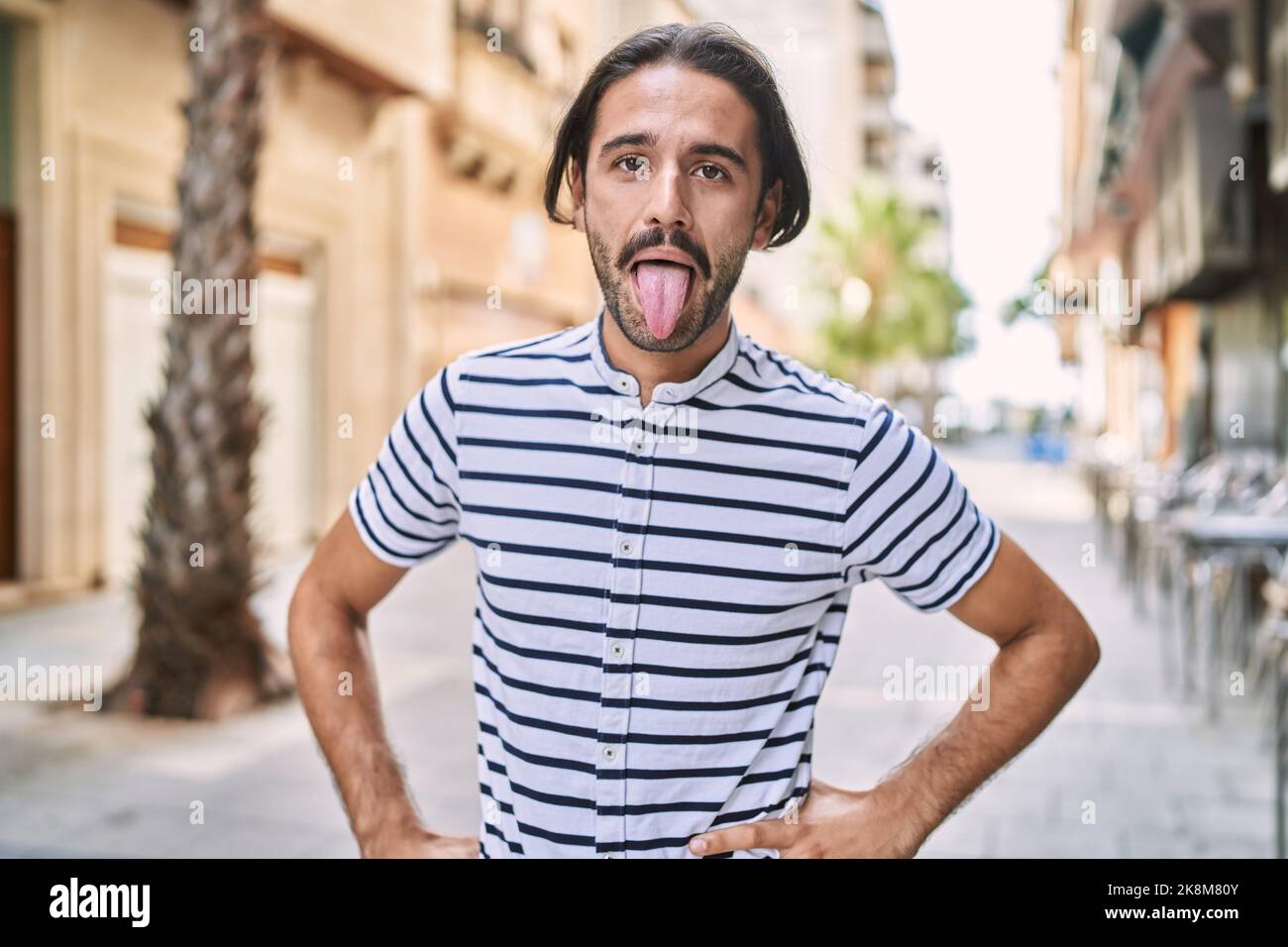 Young hispanic man with beard outdoors at the city sticking tongue out ...