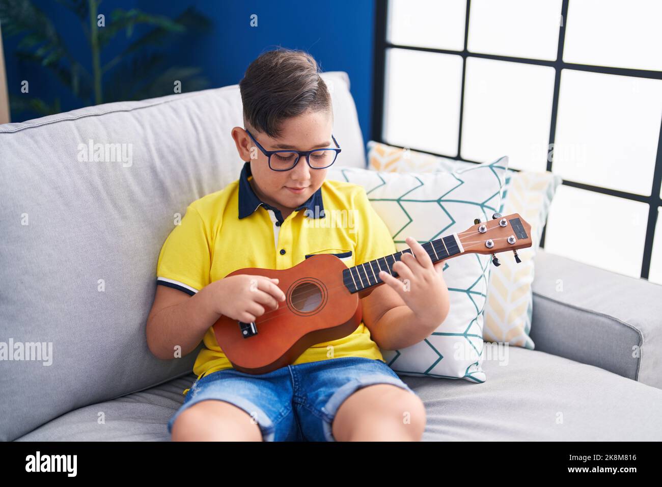 Adorable hispanic boy playing ukulele sitting on sofa at home Stock ...