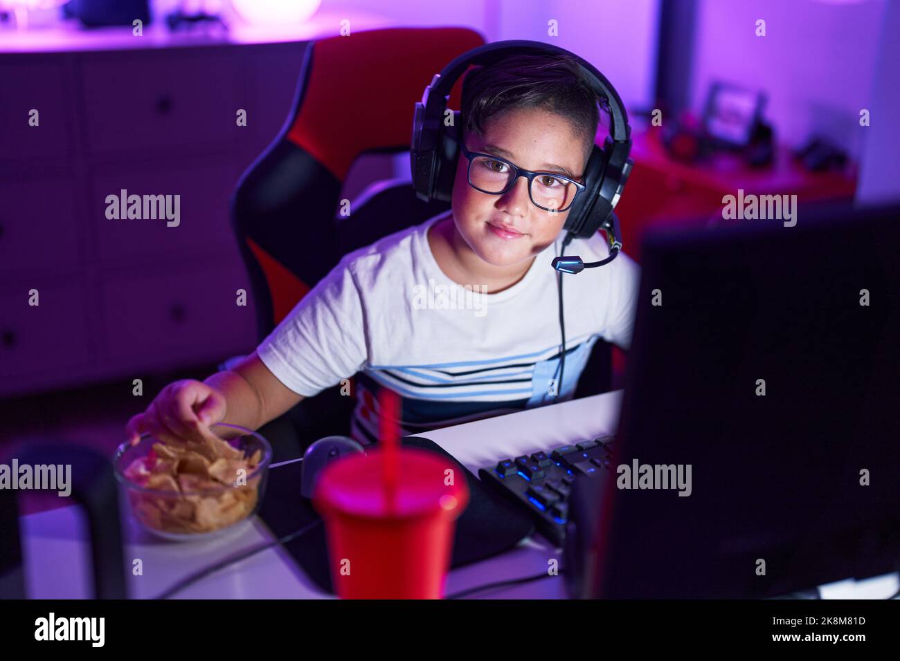 Adorable hispanic boy streamer playing video game eating chips potatoes ...