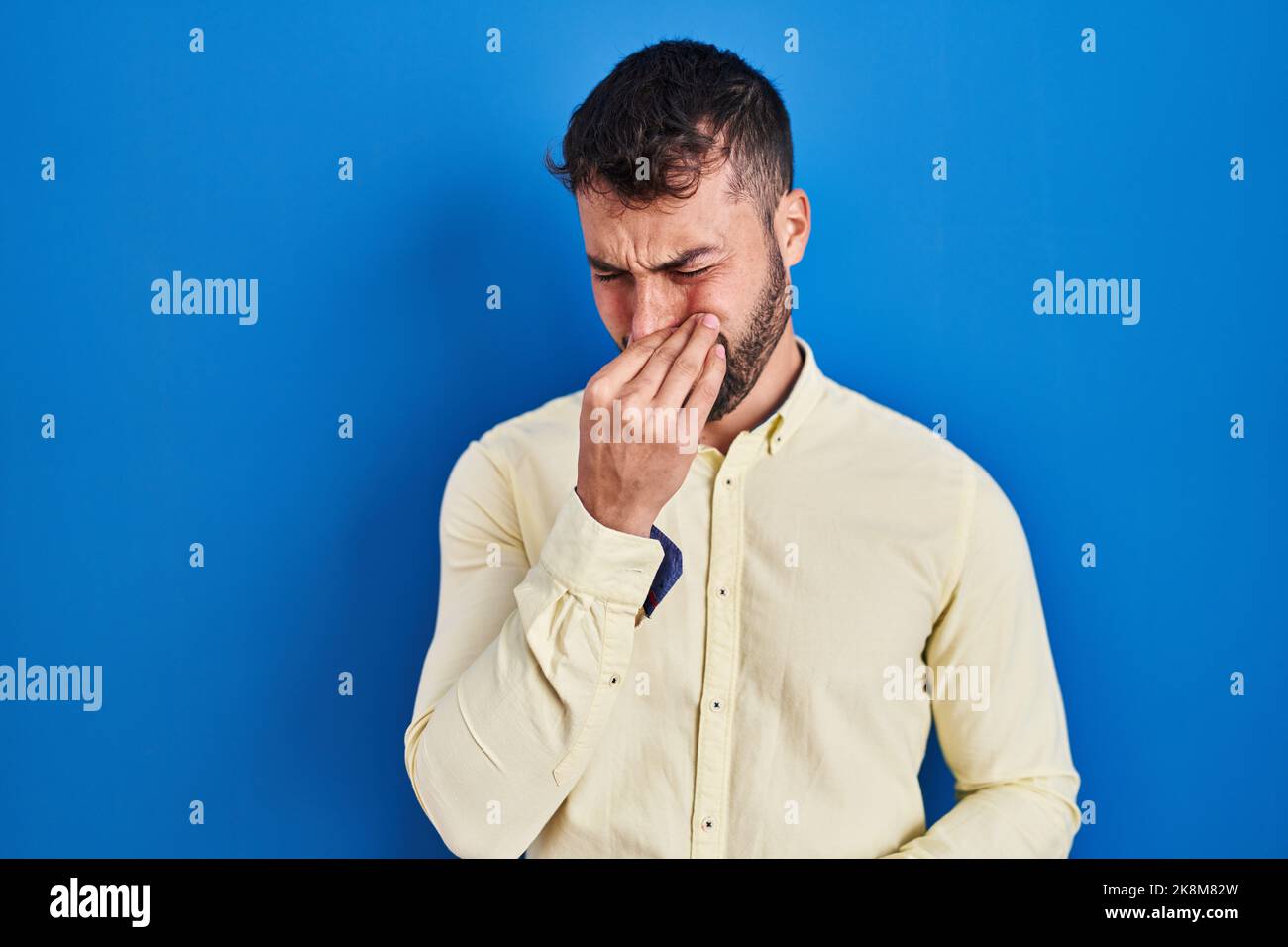 Handsome hispanic man standing over blue background smelling something ...