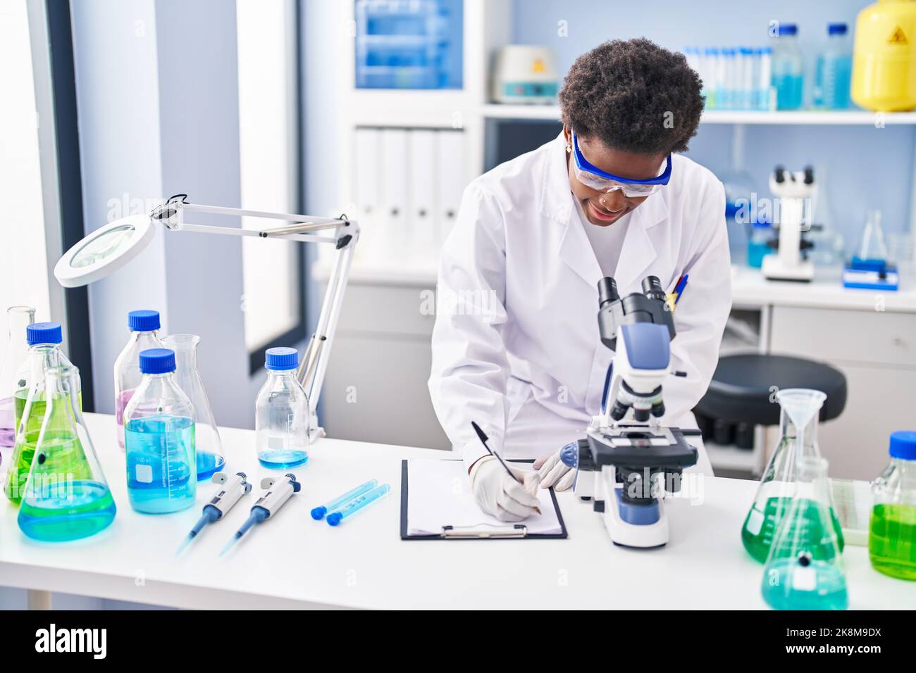 African american woman wearing scientist uniform using microscope write on clipboard at ...