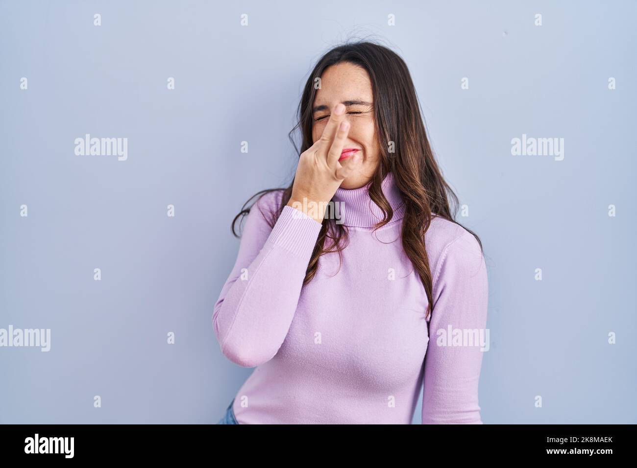 Young brunette woman standing over blue background smelling something ...