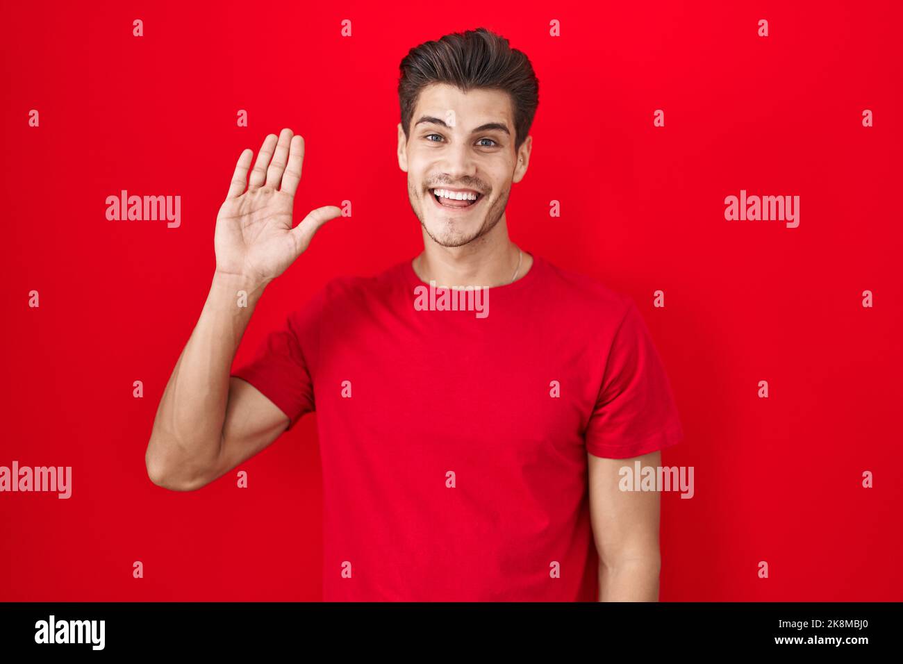 Young hispanic man standing over red background waiving saying hello ...