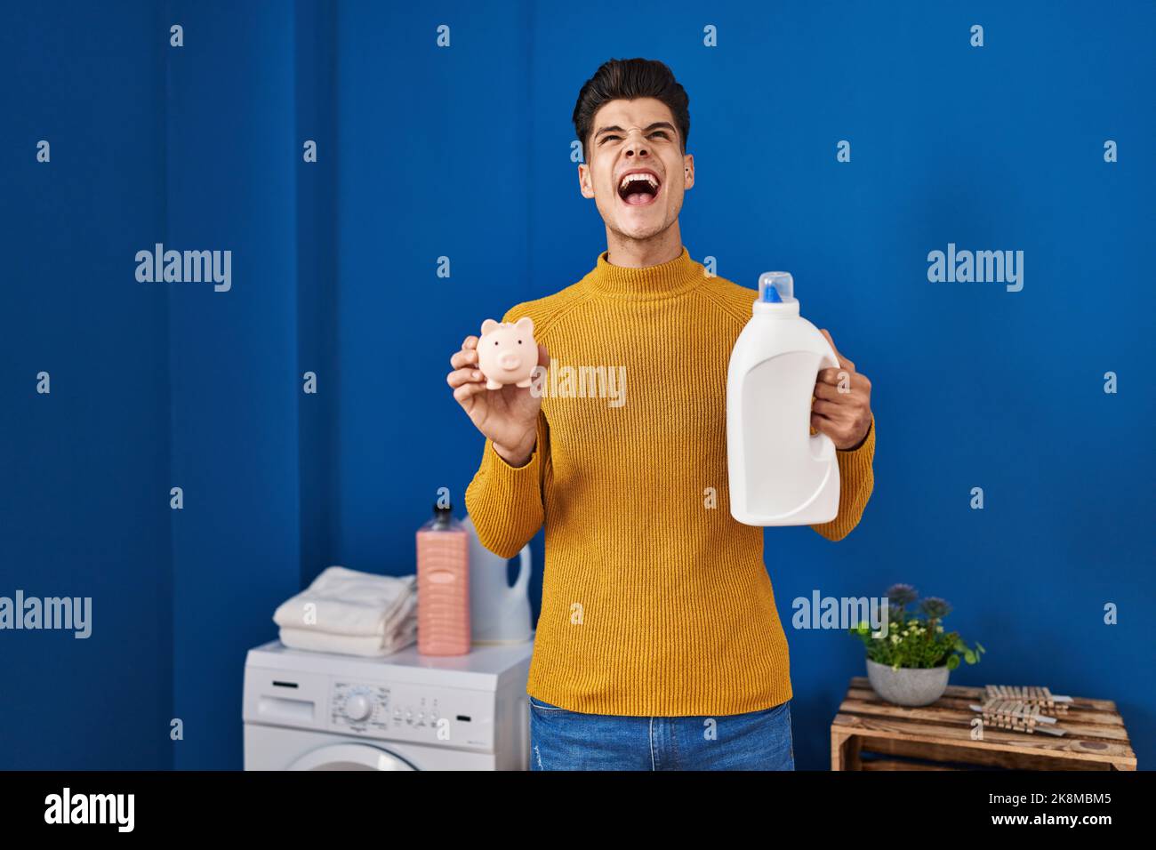 Young hispanic man holding detergent bottle and piggy bank angry and ...