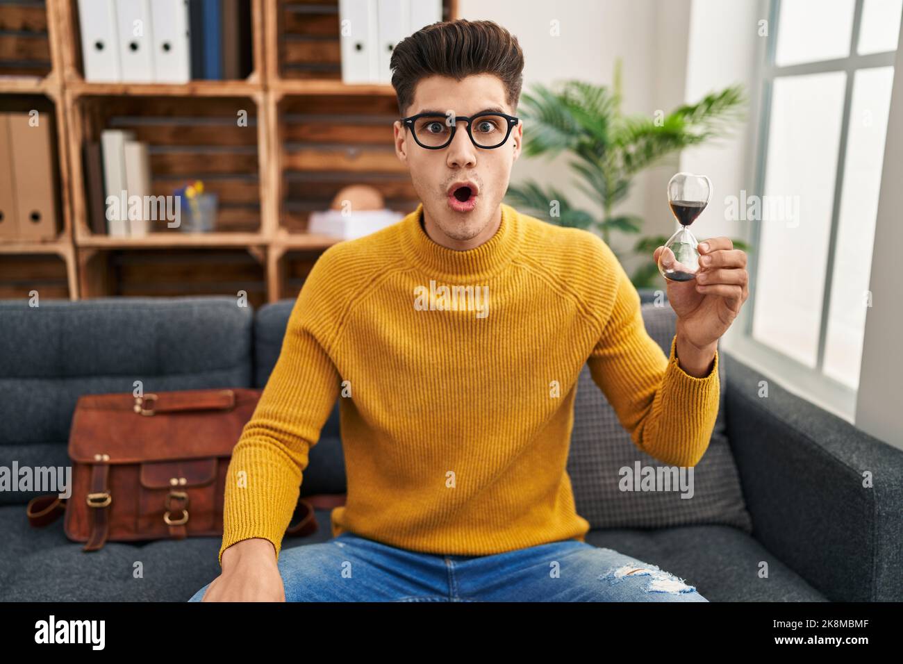 Young hispanic man holding sand clock scared and amazed with open mouth ...