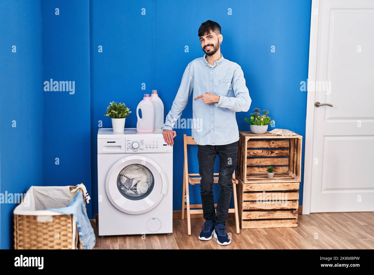 Young hispanic man with beard doing laundry standing at laundry room ...