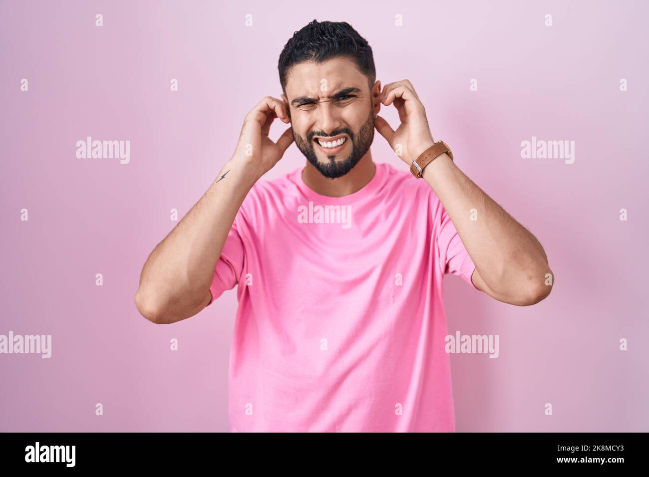 Hispanic young man standing over pink background covering ears with ...