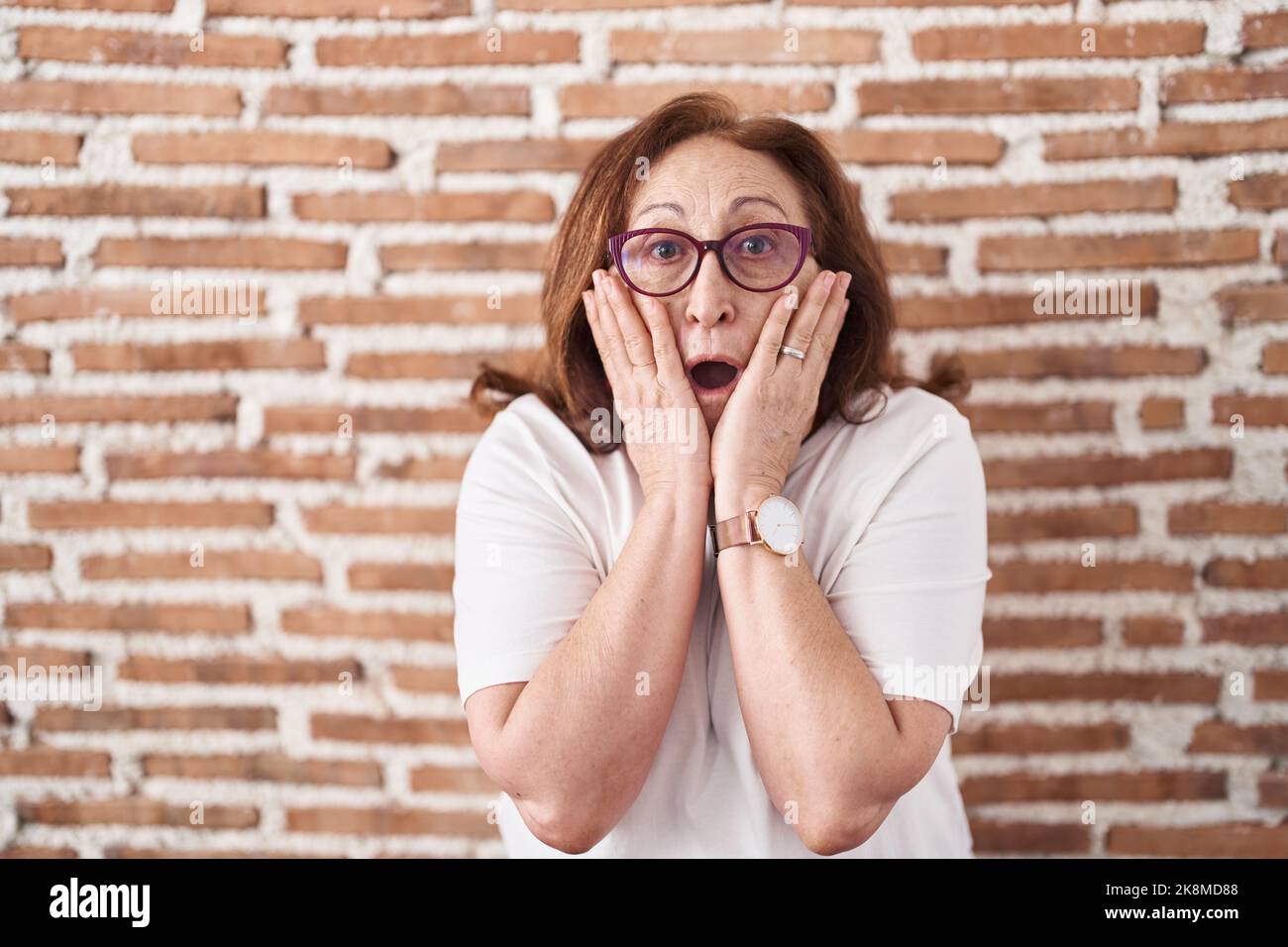 Senior woman with glasses standing over bricks wall afraid and shocked ...