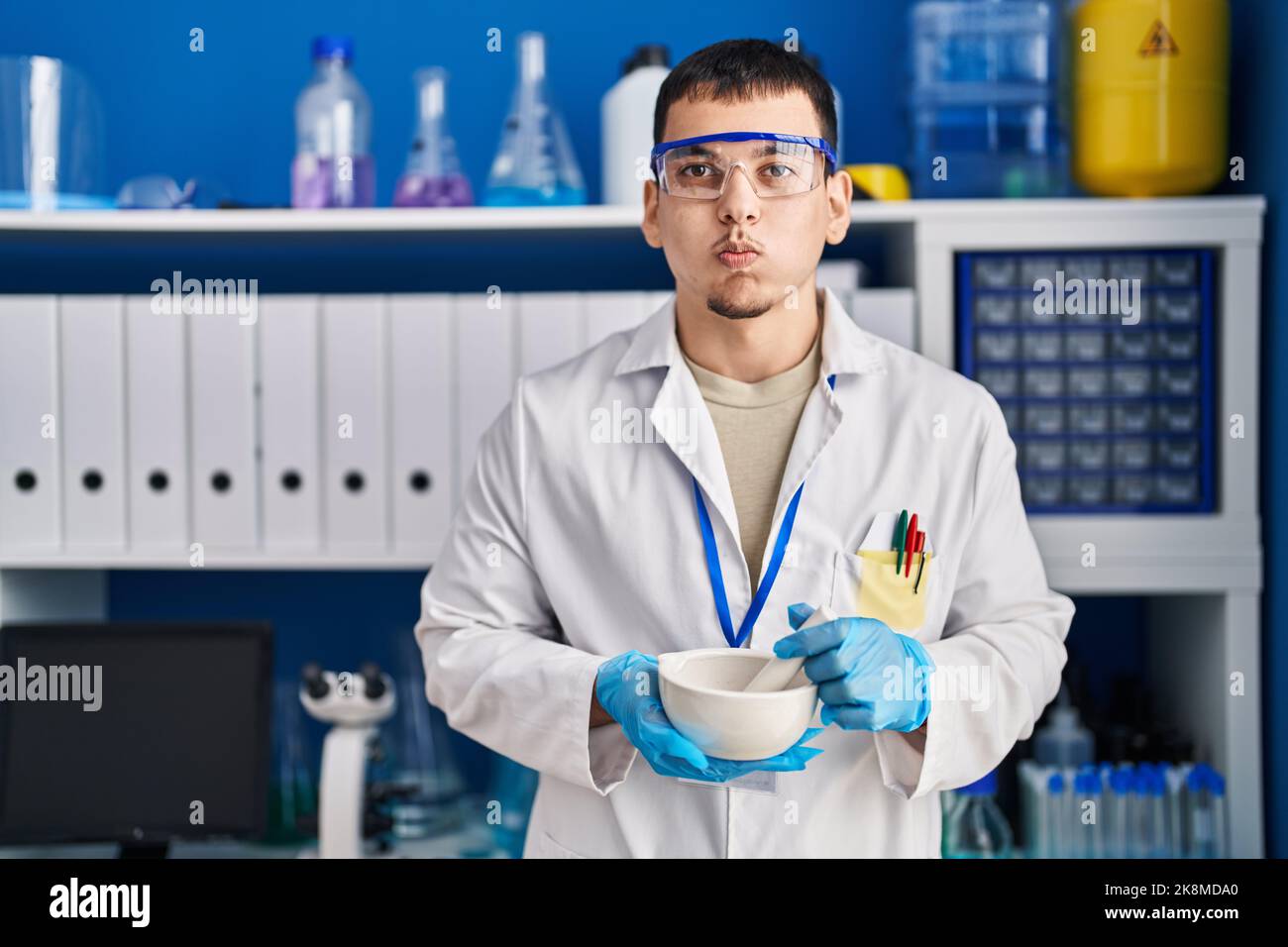 Young arab man working at scientist laboratory puffing cheeks with funny face. mouth inflated ...