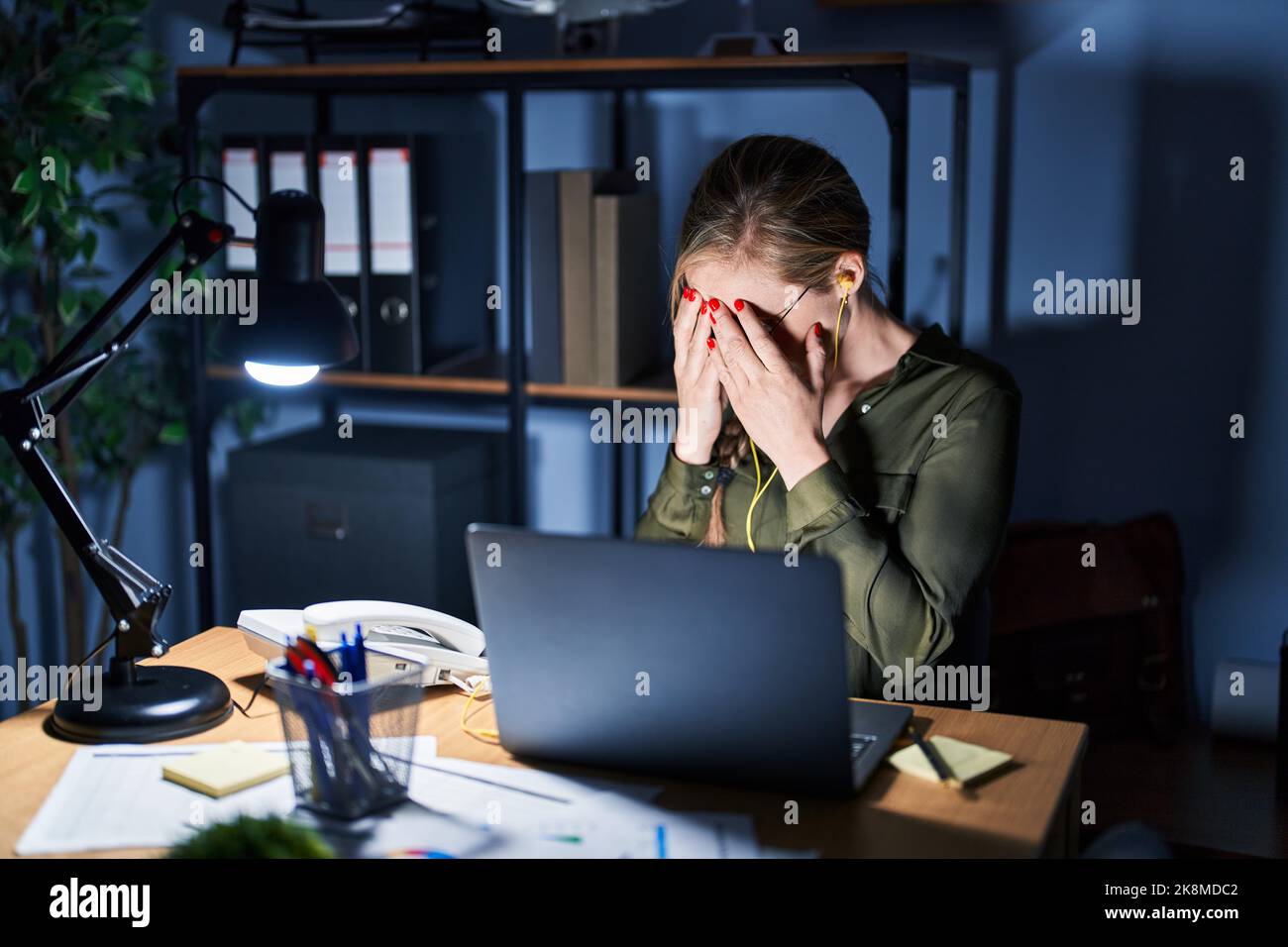Young blonde woman working at the office at night with sad expression ...