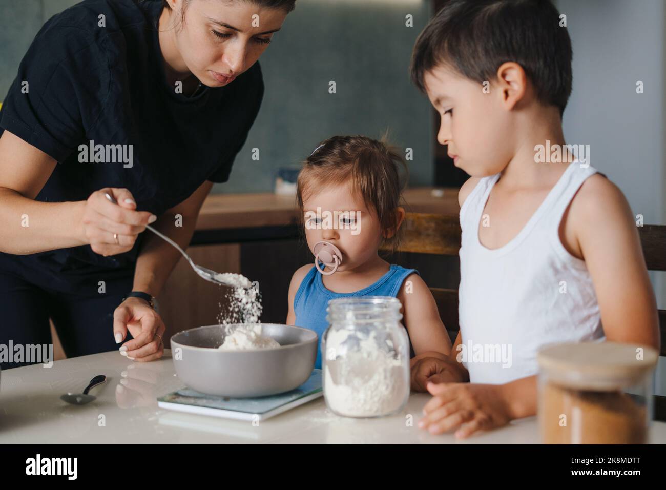 young mother with her two kids weighing out flour into bowl in kitchen ...