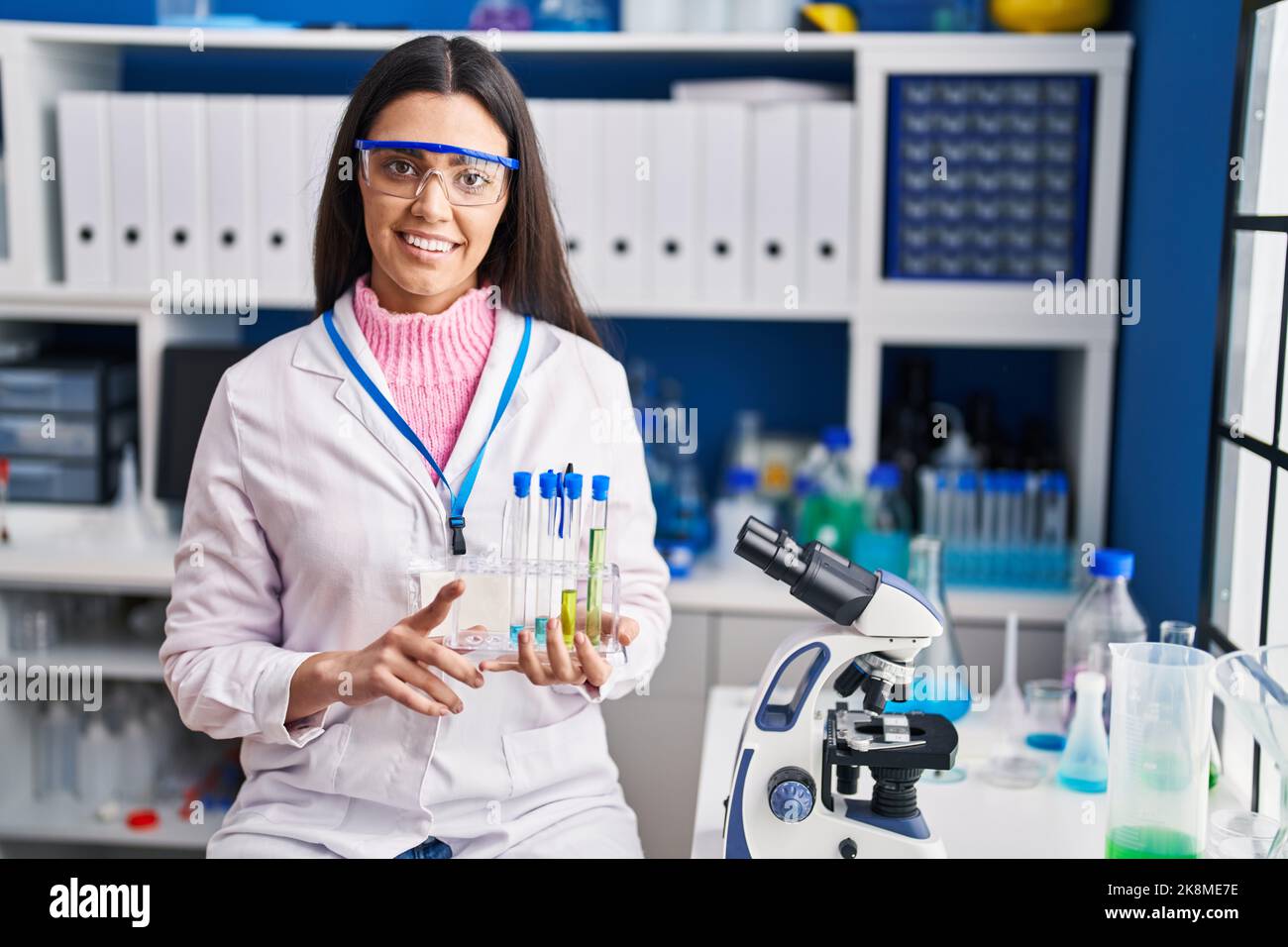 Young brunette woman working at scientist laboratory looking positive and happy standing and ...