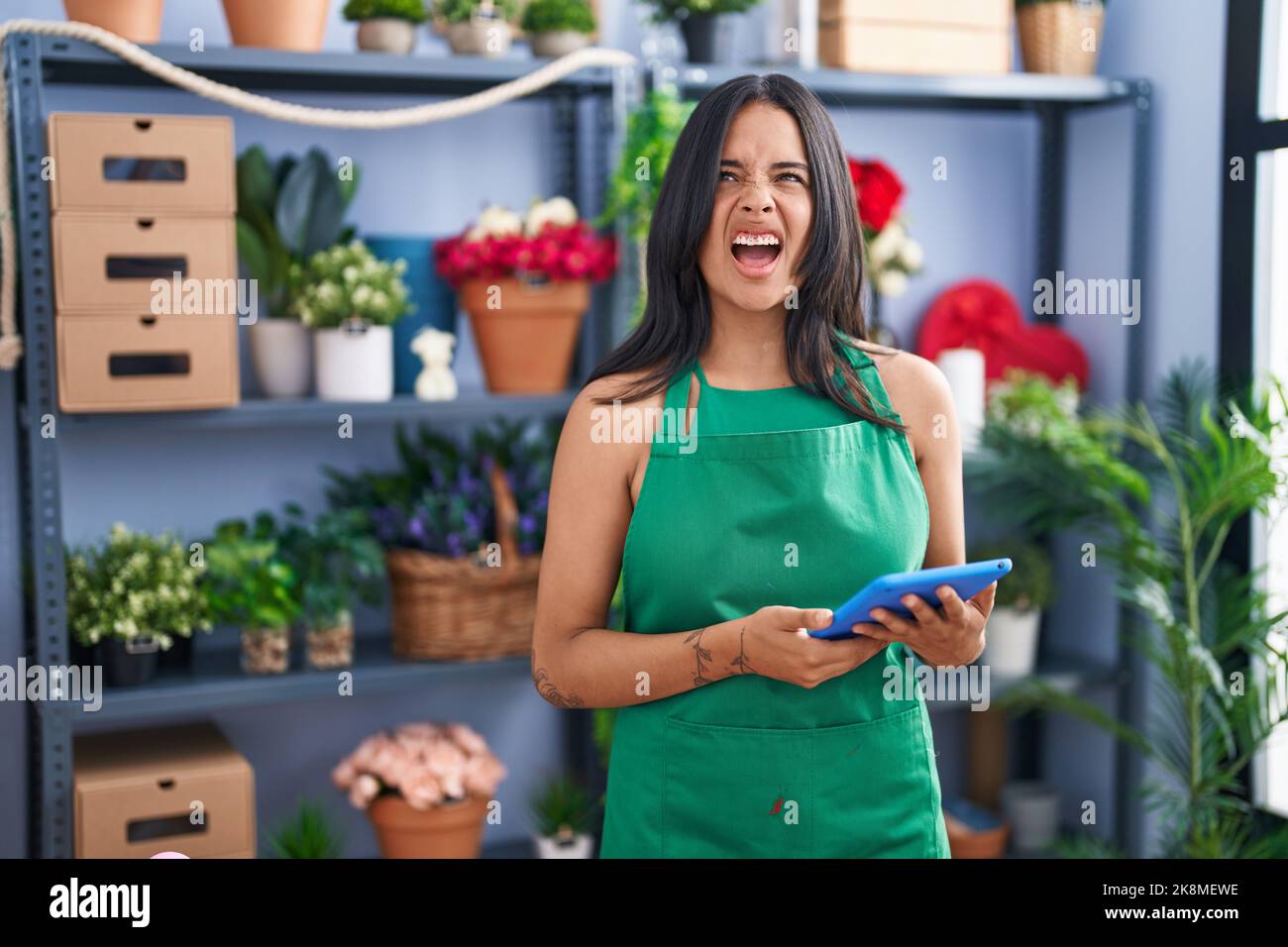 Brunette woman working at florist shop holding tablet angry and mad ...
