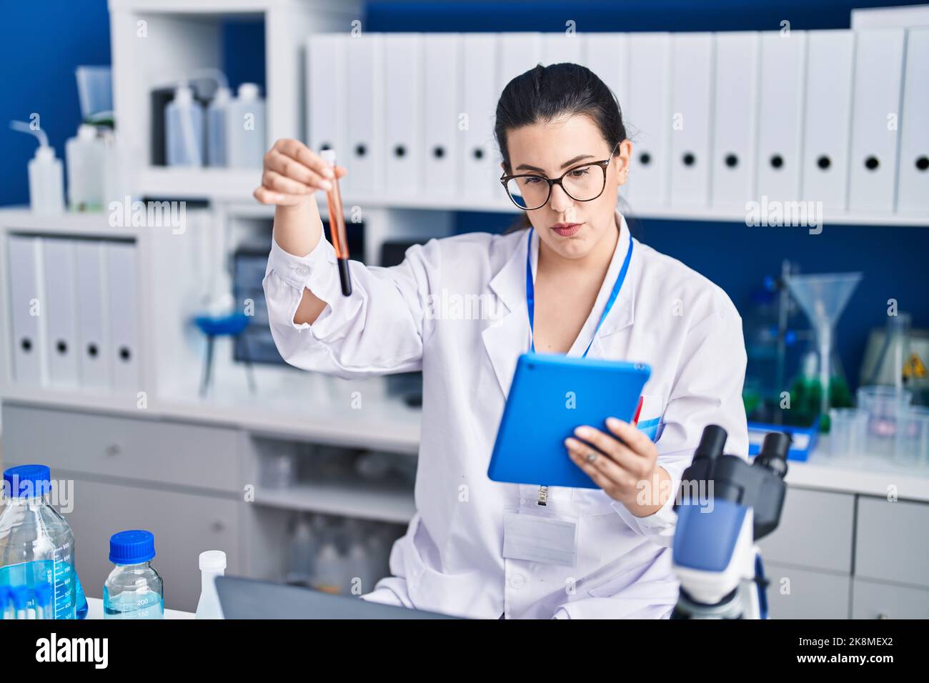 Young hispanic woman scientist using touchpad holding blood test tube ...