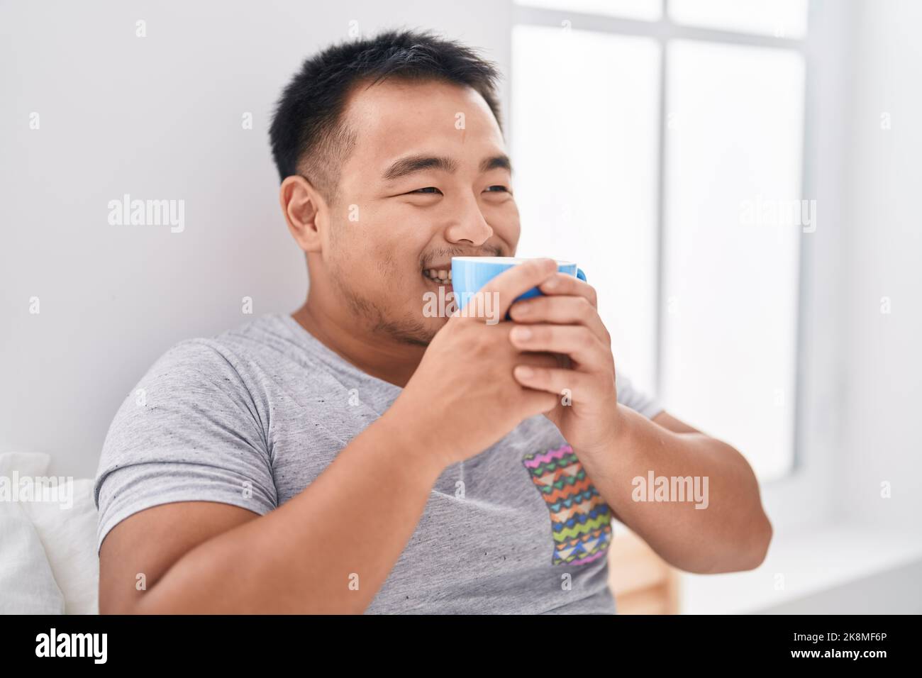 Young chinese man drinking cup of coffee sitting on bed at bedroom ...