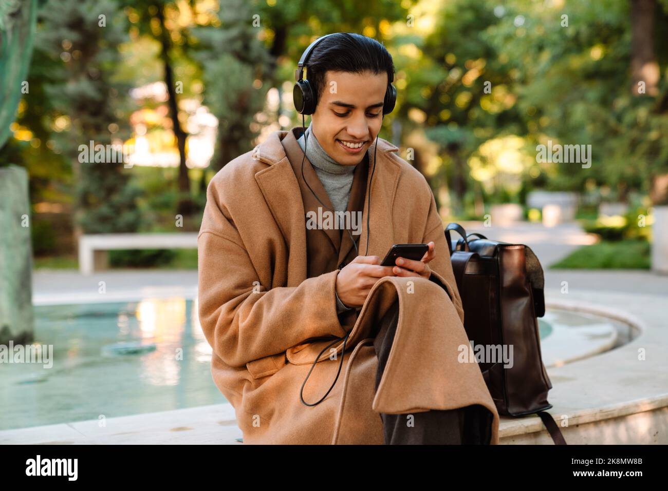 Young man in headphones smiling and using mobile phone while resting in ...