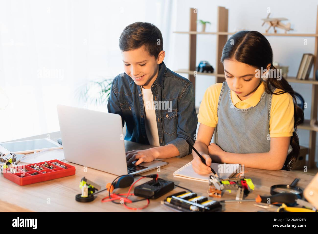 happy boy using laptop while girl writing in notebook near details of ...