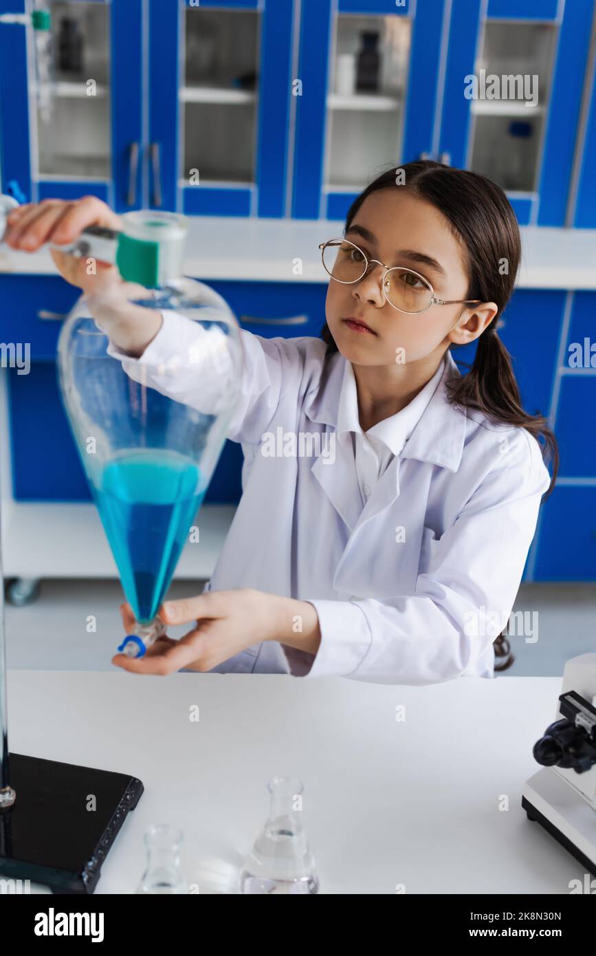 girl in eyeglasses and white coat holding flask with blue liquid in lab ...