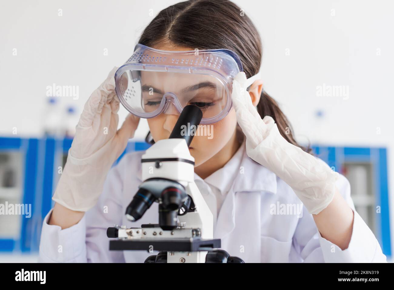 preteen girl in latex gloves and goggles using microscope during ...