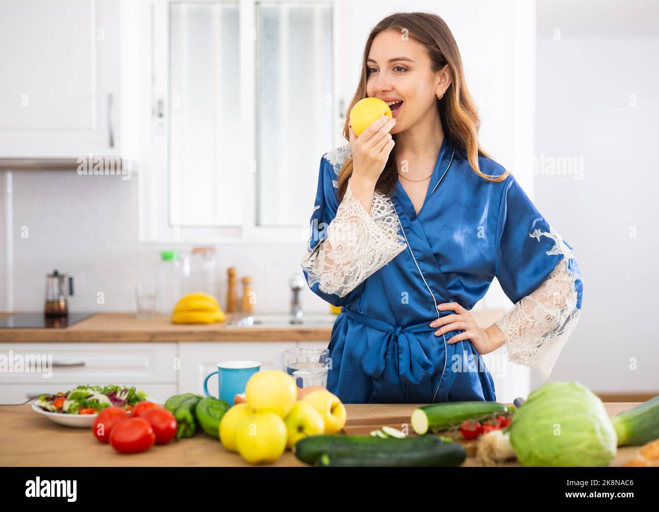 Satisfied woman in dressing gown eating apples in kitchen during ...