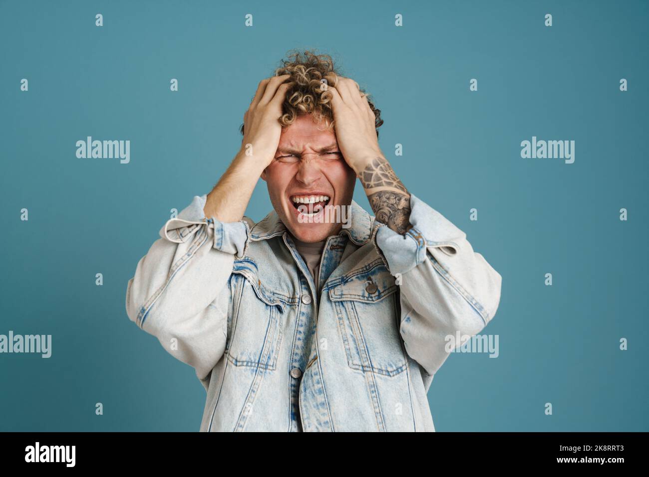 Young curly man with headache screaming and holding his head isolated ...