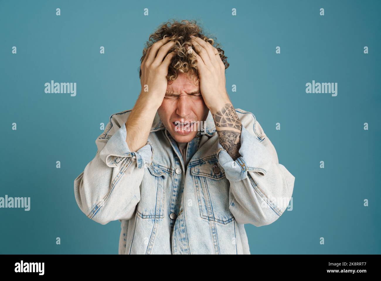Young curly man with headache screaming and holding his head isolated ...