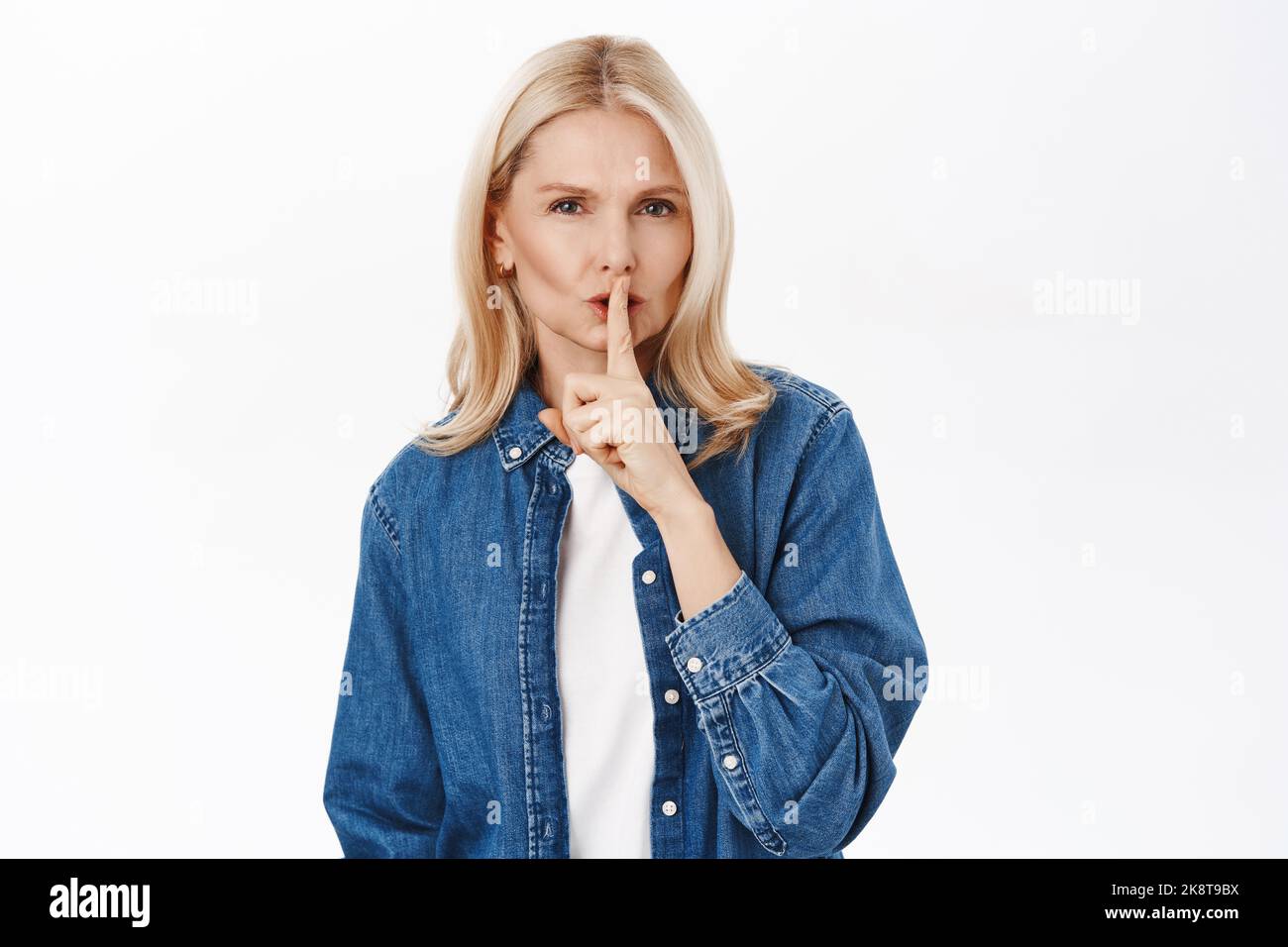 Portrait of blond senior woman shushing, press shh index finger on lips ...