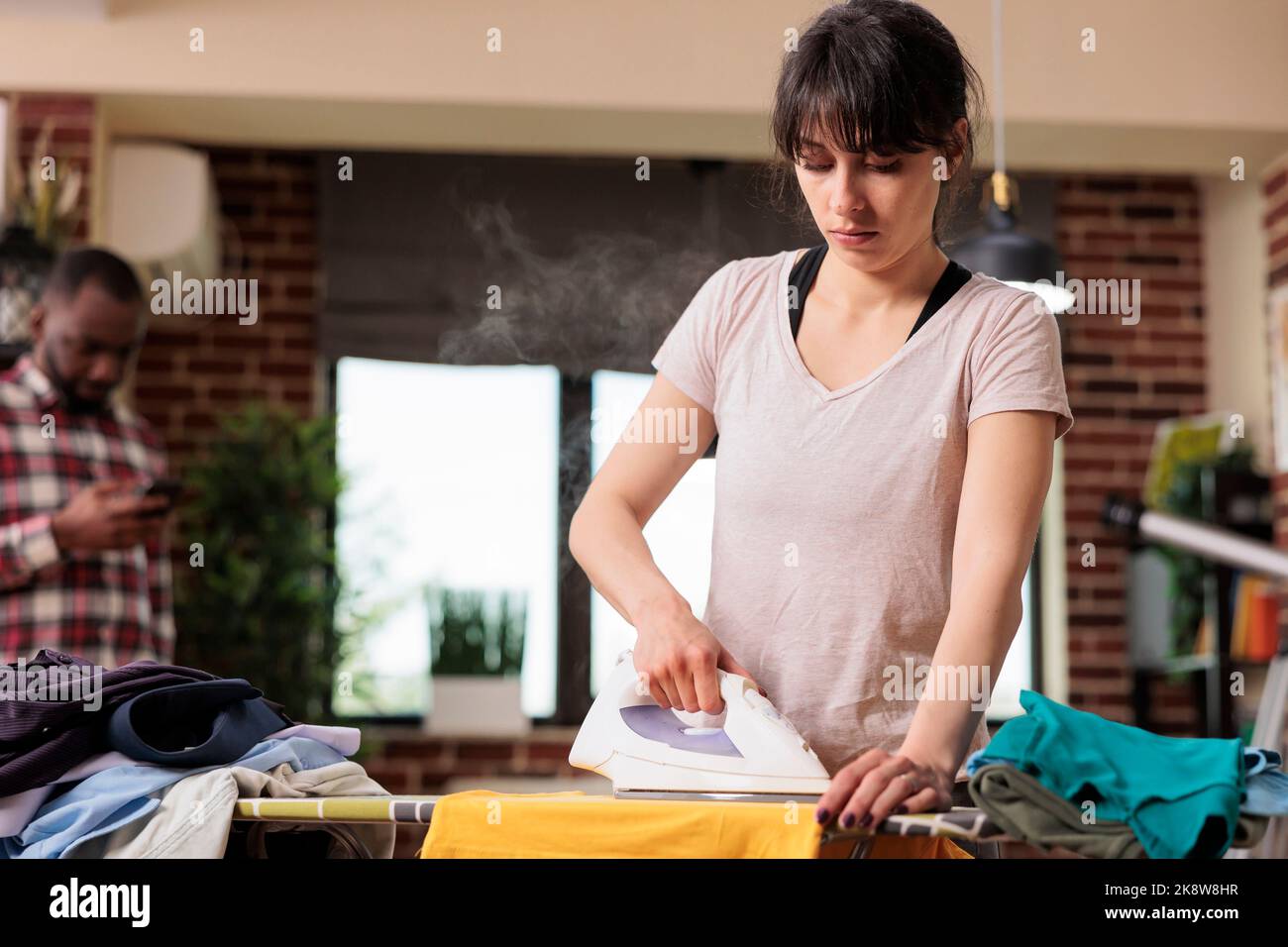 Serious woman concentrating on ironing clothes at home while boyfriend ...