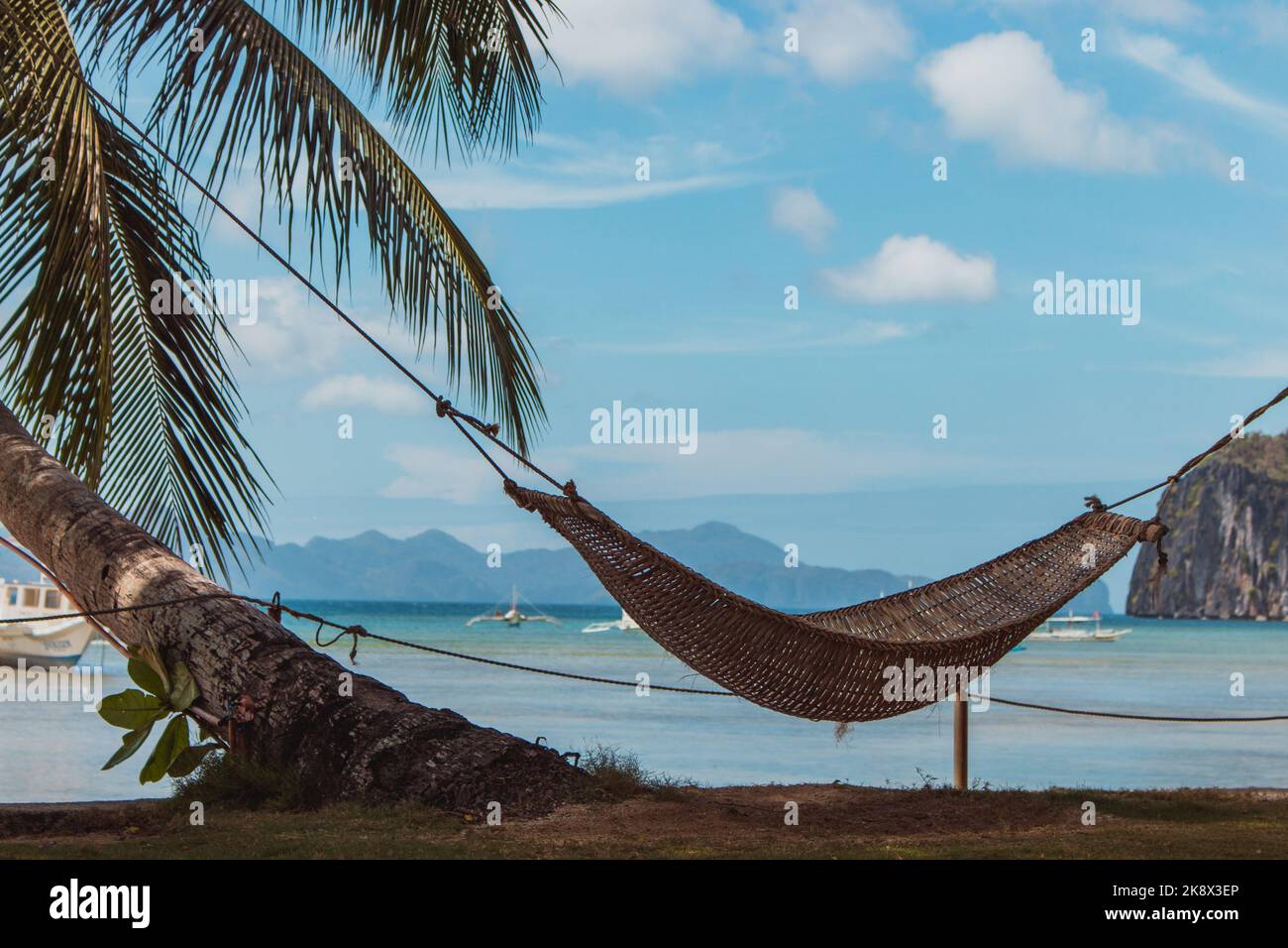 Empty hammock with palm tree and boat on tropical beach. Philippines resort landscape. Idyllic ...