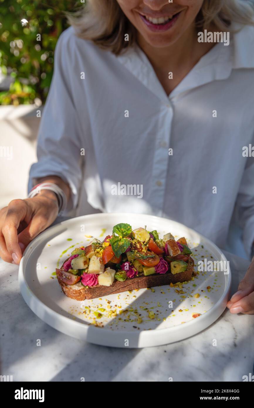Woman in white shirt with a plate of avocado, tomato and halloumi toast ...