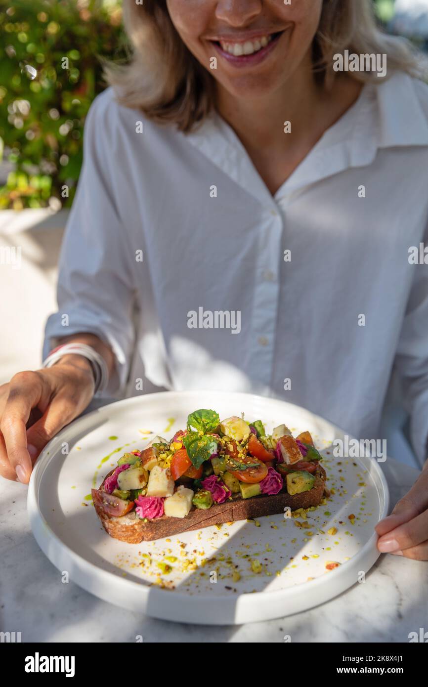 Woman in white shirt with a plate of avocado, tomato and halloumi toast ...