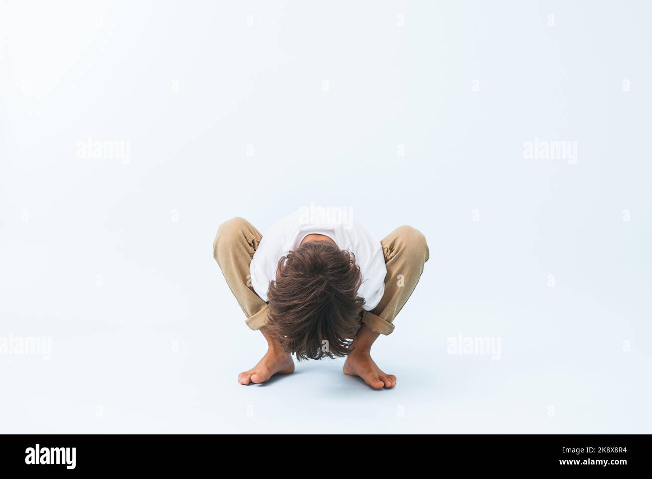 Crouching Indian boy lowering his body betweeen legs against bluish ...