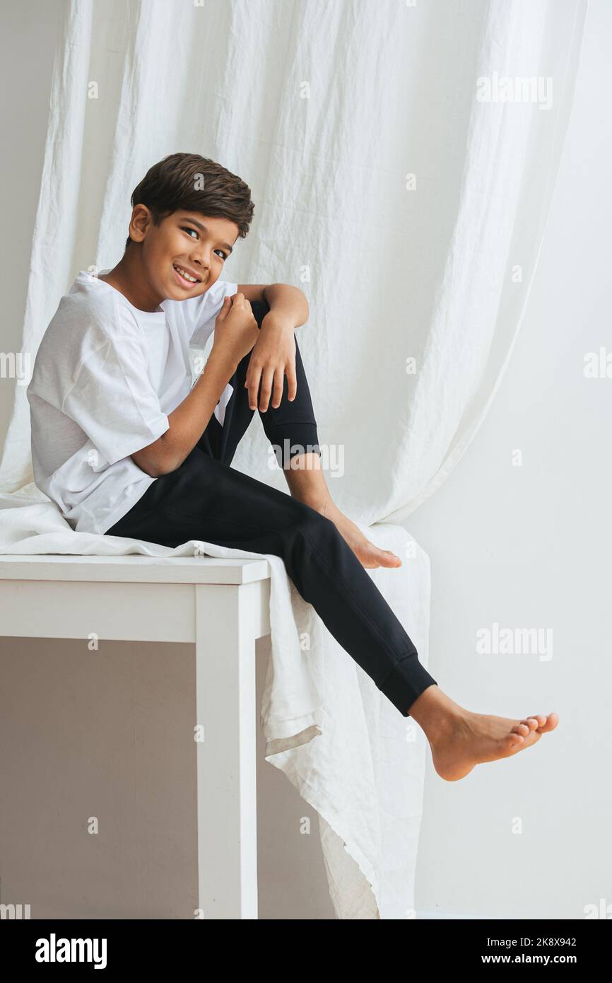 Happy indian boy sitting on a table, awkwardly smiling at the camera ...
