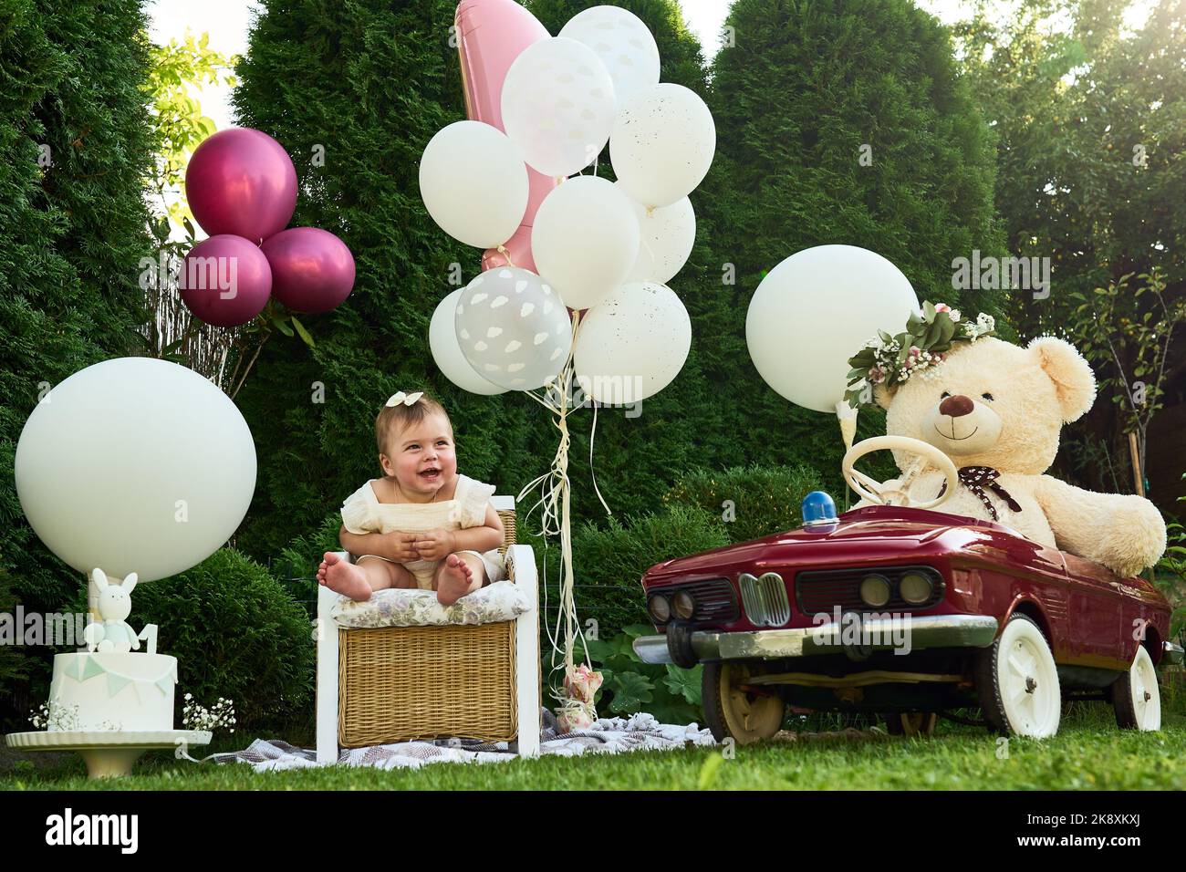 A 1-year baby girl sitting outdoors on a wooden chair with a balloons ...