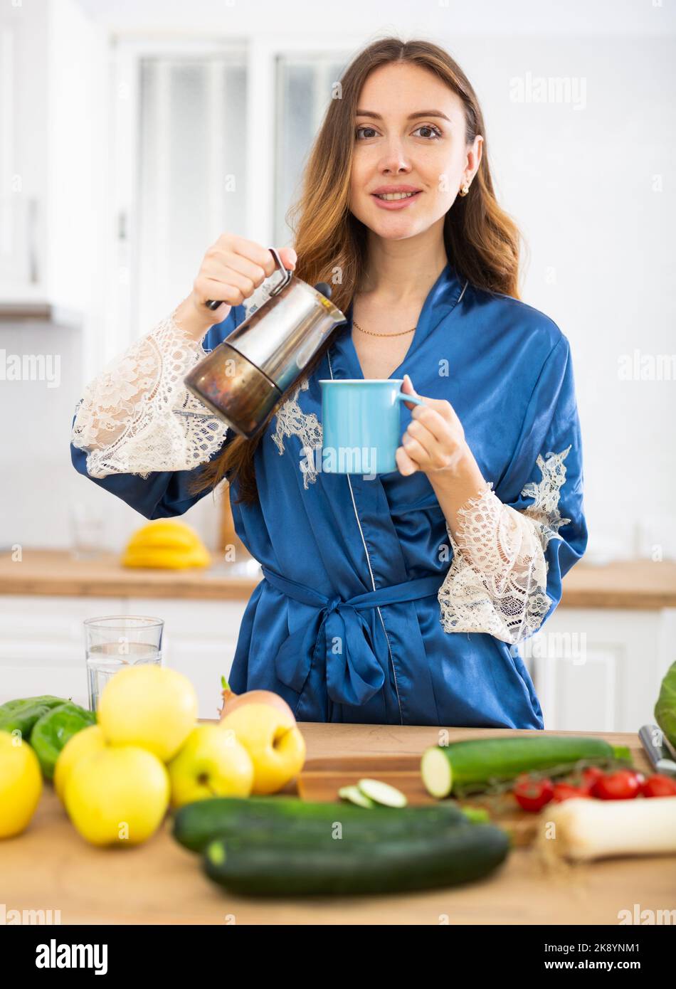 Smiling young woman in blue robe pouring coffee into cup at home ...