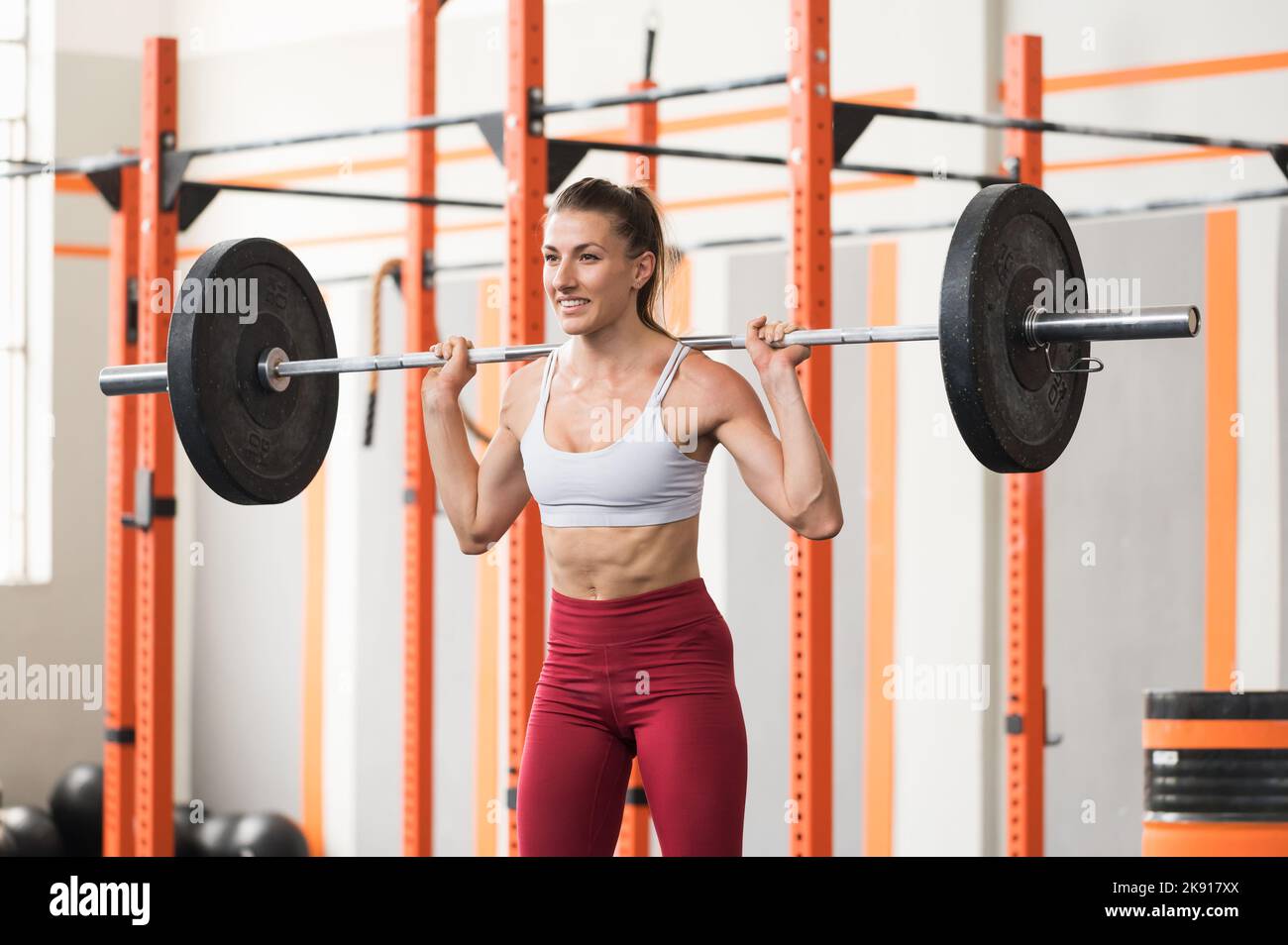 Strong young female athlete in activewear with barbell on shoulders ...