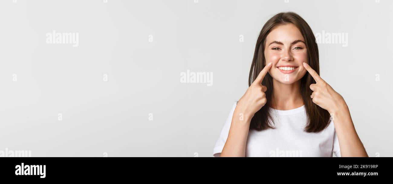 Close-up of attractive smiling young girl pointing fingers at face ...