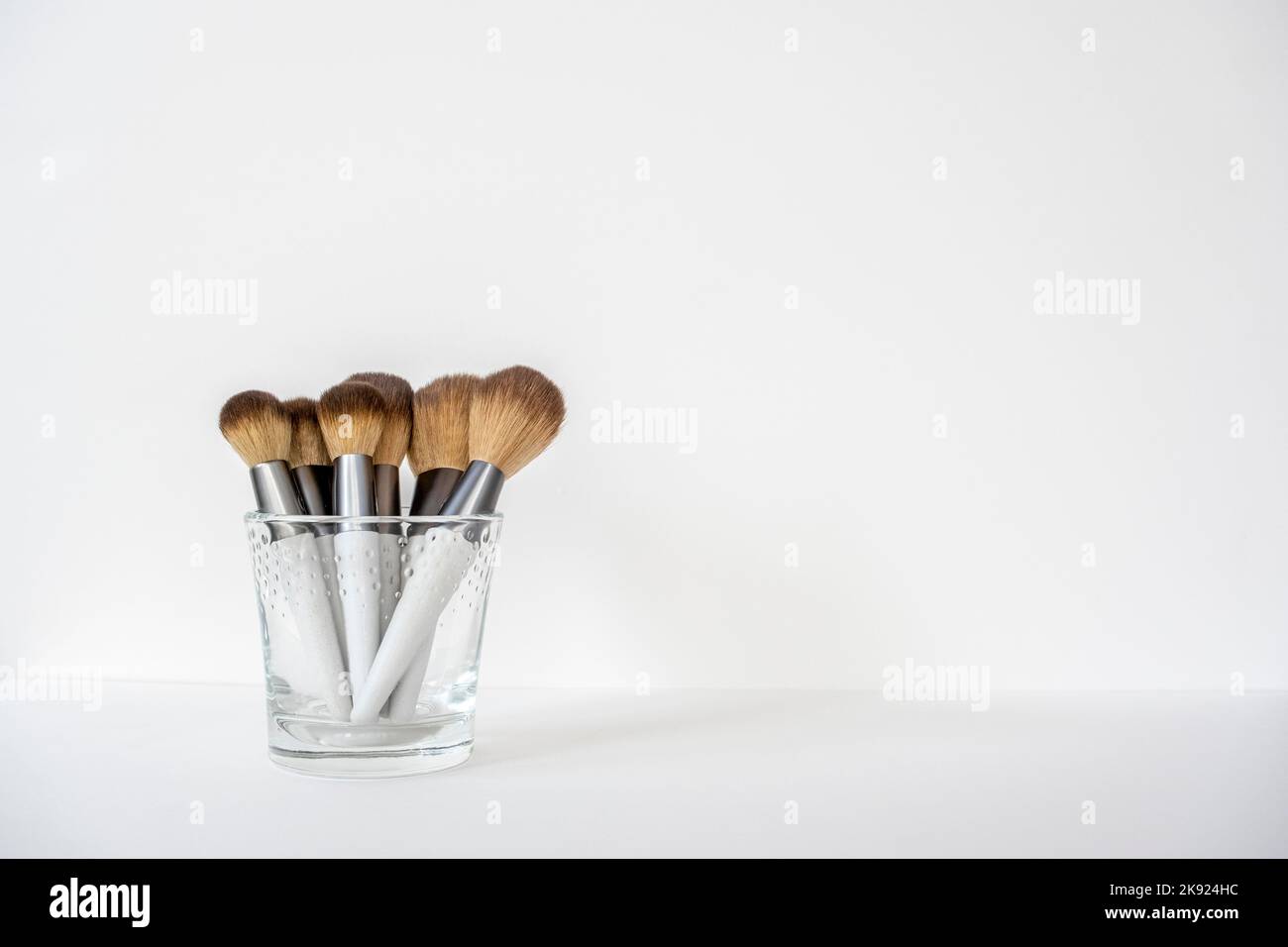 Glass cup holds makeup brushes on a white table with white background ...